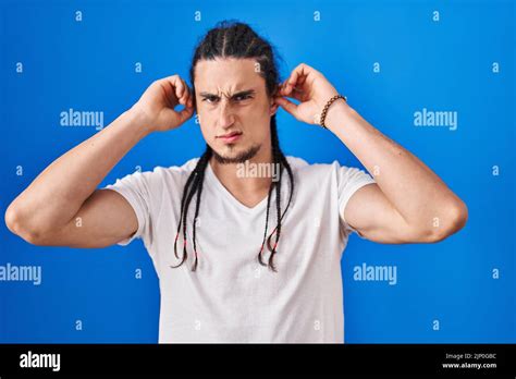 Hispanic Man With Long Hair Standing Over Blue Background Covering Ears