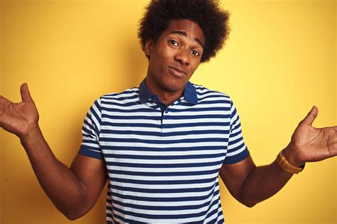 고립 된 노란색 배경 에 서 해군 줄무늬 폴로를 입고 아프로 머리를 가진 미국 남자는 팔과 손을 들고 단서와 혼란 표정을 의심 개념 감정에 대한 스톡 사진 및 기타 이미지