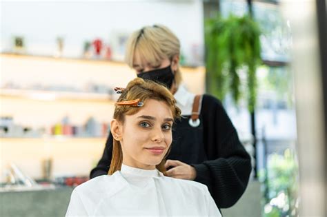 아시아 미용사는 살롱에서 아름다운 여성의 머리를 절단 마스크를 착용합니다 백인 고객 소녀는 가게에서 스타일리스트에서 이발을받고있는 동안 미소와 거울을보고 뷰티 이발소