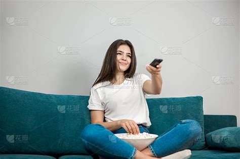 一个漂亮的年轻女子一边看电视一边吃爆米花，舒舒服服地坐在家里的沙发上素材 花瓣网