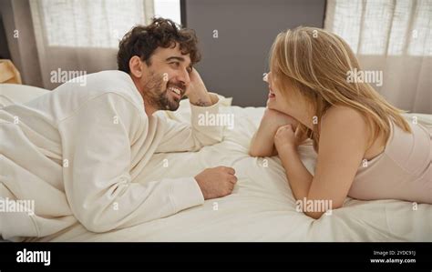 A Smiling Man And Woman Lying On A Bed Facing Each Other In A Cozy Bedroom Depicting A Loving