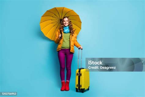 예쁜 여행자 레이디 홀드 우산 롤링 백 가방 대기 공항 여행 착용 노란색 오버 코트 블루 스카프 바이올렛 바지 부츠 고립 된 블루 컬러 배경의 전체 길이 사진 파란색 배경에