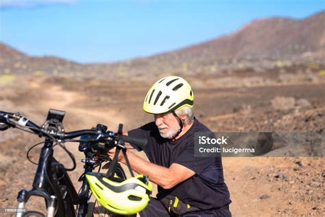 흰 머리와 수염을 가진 노인이 검은 색 전기 자전거의 배터리를 확인합니다 야외 보도 노란색 헬멧 거리가 먼에 대한 스톡 사진 및