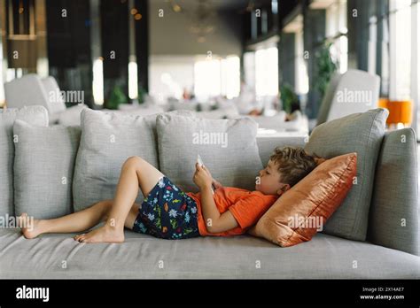 A Boy With Curly Hair Is Reclining Comfortably On A Grey Sofa With
