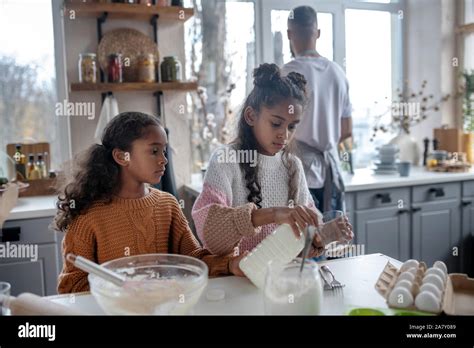 Sister Pouring Milk Into Glass While Cooking Pie With Sister Stock