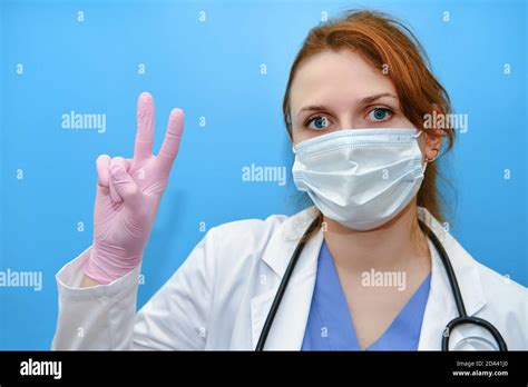 Female Doctor Shows A Gesture With His Hands Redhead Woman In Medical Mask On A Blue Background