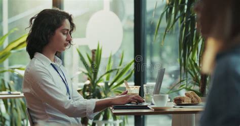Young Woman Gets Distracted From Laptop Work By Female Colleague Talking In Office Cafeteria