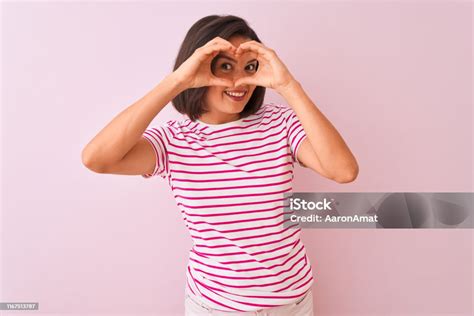 줄무늬 티셔츠를 입은 젊은 아름다운 여성은 사인을 통해 보고 미소 와 손과 손가락으로 심장 모양을하고 고립 된 분홍색 배경 위에 서 눈 신체 부분에 대한 스톡 사진 및 기타