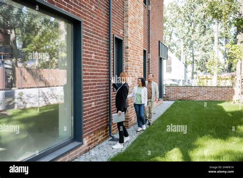 Full Length Of Redhead Realtor Holding Folder While Showing House To Happy Multiethnic Couple