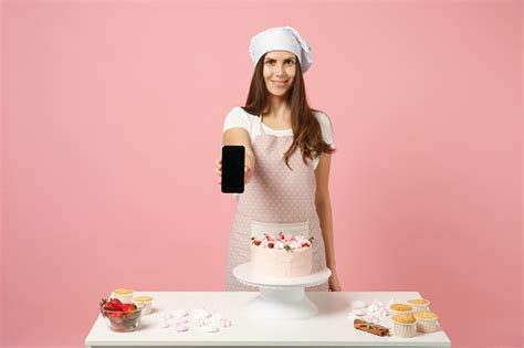 요리사 요리 과자 또는 제빵에 앞치마 흰색 티셔츠 모자 요리사 모자 요리 케이크 또는 테이블에서 컵 케 잌은 스튜디오에서 분홍색 파스텔 배경에 고립 된 핸드폰을 유지 합니다