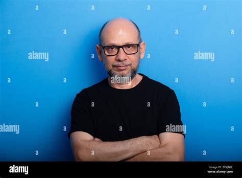 Bearded Bald Man Wearing Prescription Glasses With Arms Crossed Posing For Photo Against Blue