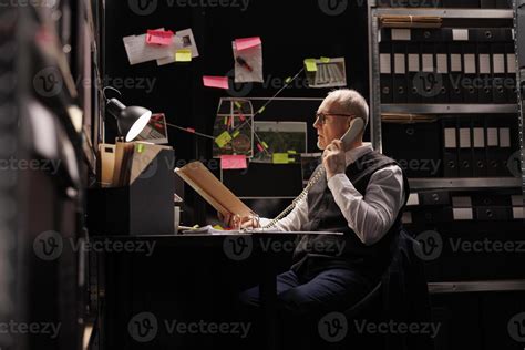 Elderly Police Officer Analyzing Criminal Case Files Evidence While Talking At Landline Phone