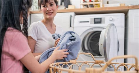 아시아의 행복한 가족은 집에 머물고 어머니는 딸에게 더러운 옷을 씻도록 가르칩니다 어린 귀여운 소녀 아이는 집에서 세탁기에 세탁물을 넣는 것을 부모 엄마로부터 돕고