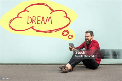 바닥에 앉아있는 빨간 셔츠에 행복 한 수염 사업가 들고 그의 디지털 태블릿을 보고 꿈 꿈 텍스트와 벽에 노란색 구름 모양 건물의