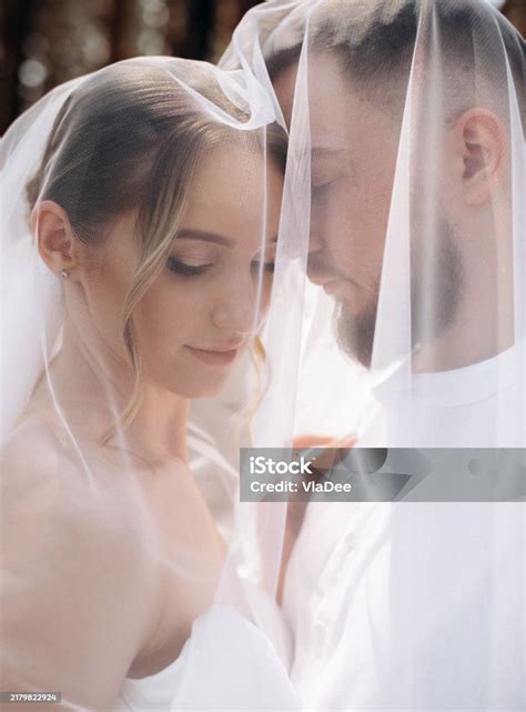 흰 셔츠를 입은 젊은 신랑과 귀여운 미소 짓는 금발 신부가 긴 베일 아래에서 껴안고 있습니다 사랑에 빠진 신혼 부부의 결혼식