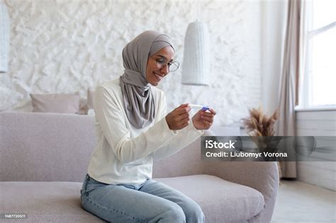 옆에서 사진 히잡을 쓴 젊은 무슬림 여성이 집 소파에 앉아 일회용의 빠른 임신 테스트기를 들고 웃고 있다 긍정적 인 결과에 만족합니다 30 34세에 대한 스톡 사진 및 기타