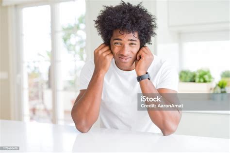 시끄러운 음악의 소음에 대한 성가신 표정으로 손가락으로 귀를 덮고 집에 앉아 캐주얼 한 흰색 티셔츠를 입은 젊은 아프리카 계 미국인 남자 청각 장애인 개념입니다 T 셔츠에