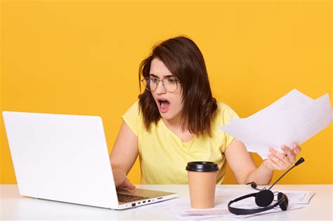 깜짝 놀란 표정으로 테이블에 앉아 있는 동안 흰색 노트북에서 일하는 젊은 아름다운 사업가의 수평 샷과 입을 열고 문서를 손에
