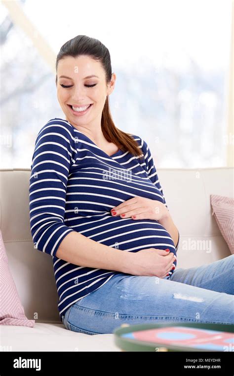 Happy pregnant woman sitting on sofa and touching her baby bump while relaxing at home Stock ...