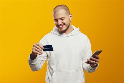 수염 난 남자는 온라인 쇼핑을 하는 신용 카드를 들고 휴대 전화를 사용하여 흰색 후드티를 입는다 지불 카드에 대한 스톡 사진 및 기타 이미지 Istock