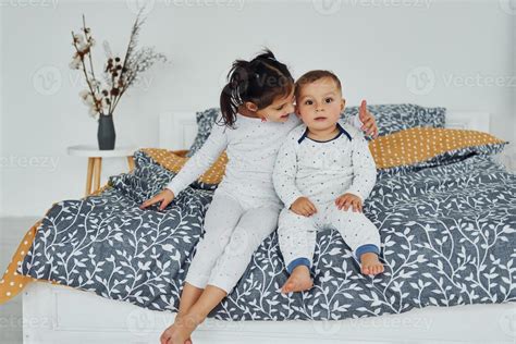 Little Sister And Brother Sits Together On The Bed Interior And Design