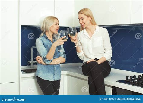 Two Blonde Girlfriends Drink Wine In The Kitchen And Laugh Two Adult