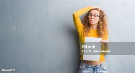 머리에 손으로 압박 하는 빈 종이 시트를 보유 하는 회색 그런 지 벽에 젊은 빨강 머리 여자는 수치심과 깜짝 얼굴로 충격 분노와 좌절 두려워 하 고 실수에 대 한 화가