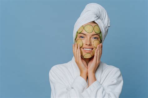 젊은 기쁘게 여자 는 얼굴에 오이 조각을 적용 하고 미용 절차는 흰색 목욕 코트와 파란색 벽에 고립 된 머리에 감싸인 수건을 입고 피부를 젊어지게하는 얼굴 마스크를 만듭니다