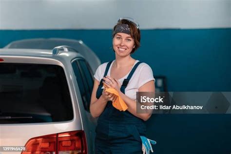 푸른 작업복을 입은 행복한 백인 젊은 여성이 헝겊으로 손을 흔들고 있는 모습 배경에있는 자동차와 워크샵 자동차 수리점에서 일하기