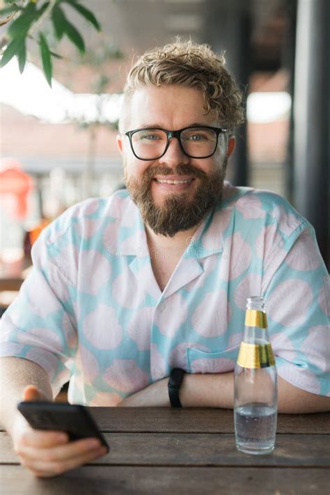 Millennial Cheerful Man With Glasses And A Patterned Shirt Smiles Warmly While Sitting At An